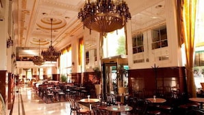 Um amplo hall de hotel com lustres elaborados, diversas áreas de estar e grandes janelas que proporcionam luz natural.