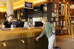 Um balcão de auto check-in com uma placa acima, uma área de bar com garrafas e uma estante de livros com livros e uma escada.