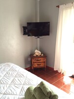 Quarto com cama, televisão fixada na parede, mesa de cabeceira de madeira e janela com cortinas.