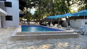 Uma piscina com ladrilhos de pedra, rodeada por espreguiçadeiras brancas e guarda-sóis azuis.