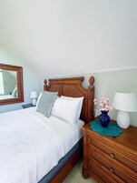 Cama bem arrumada com lençóis brancos e cabeceira de madeira, mesa de cabeceira com um vaso de flores e um abajur.