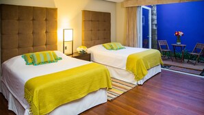 Duas camas de casal com colchas listradas em amarelo e verde, chão de madeira e uma pequena mesa com cadeiras em um quarto com parede azul.