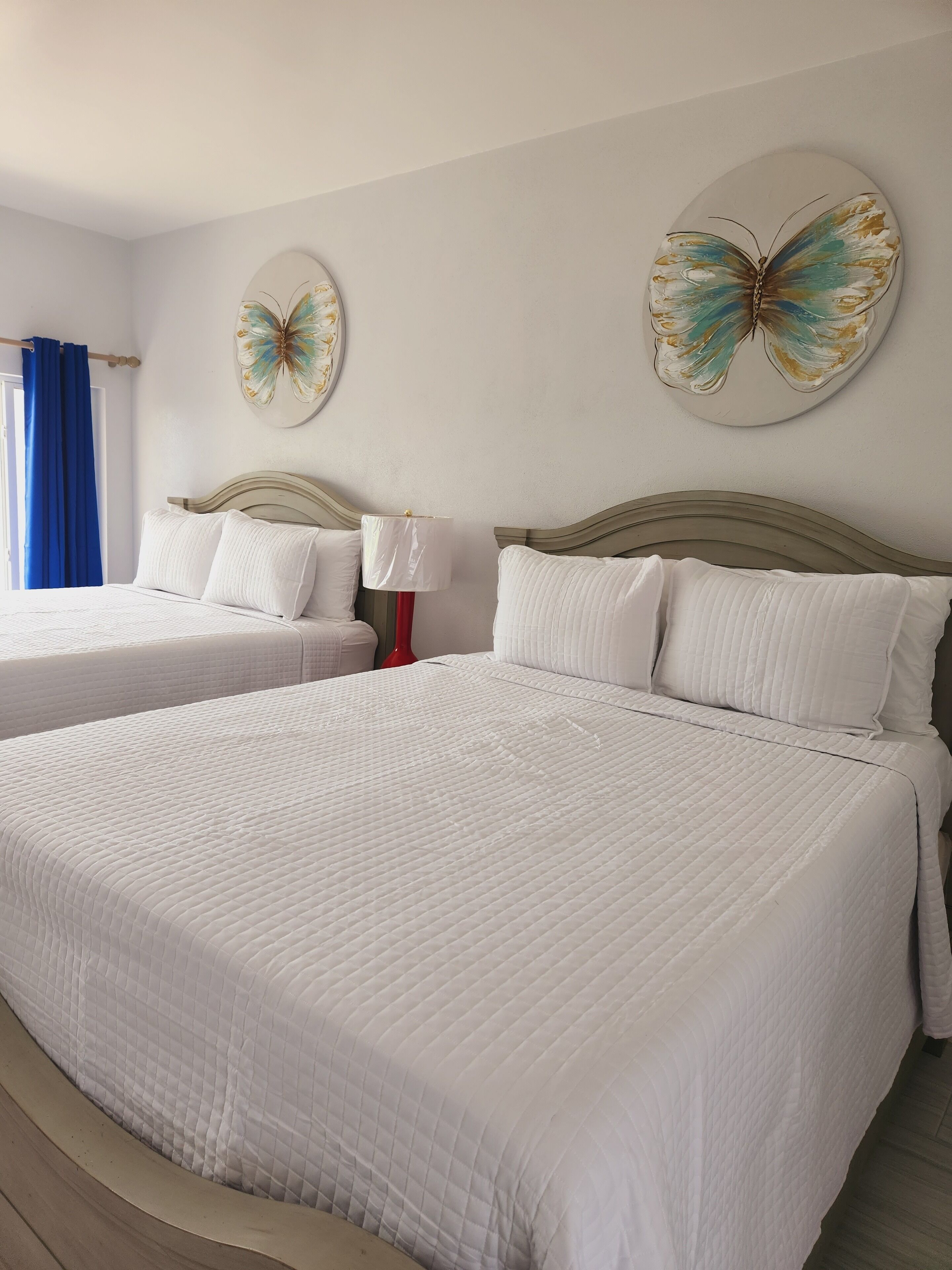 Quarto de hotel com duas camas, roupa de cama branca, abajur vermelho e arte de parede com borboletas.