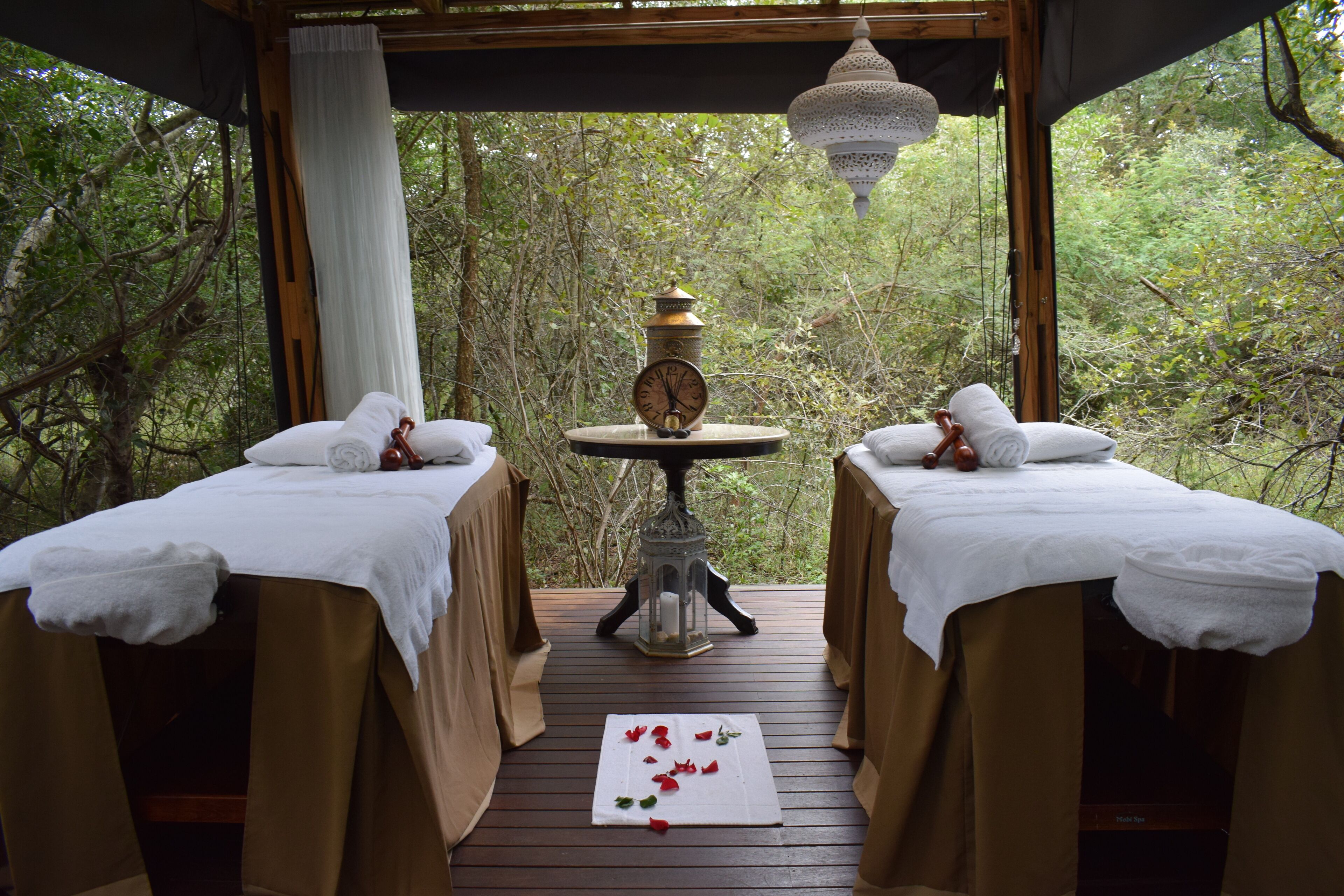 Um spa tranquilo com duas mesas de massagem, um candeeiro decorativo e um deck de madeira rodeado por vegetação exuberante.