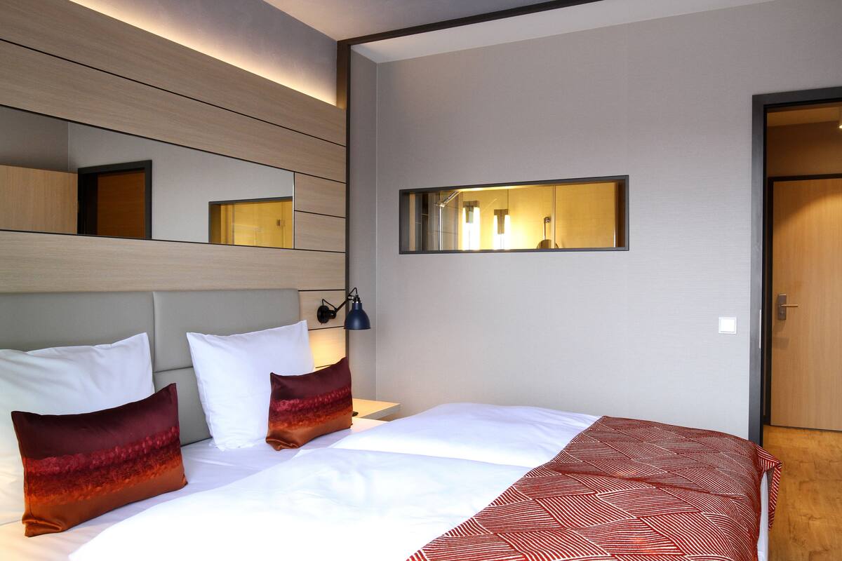 Quarto de hotel com uma cama grande, cabeceira, mesa de cabeceira com abajur e um espelho na parede.