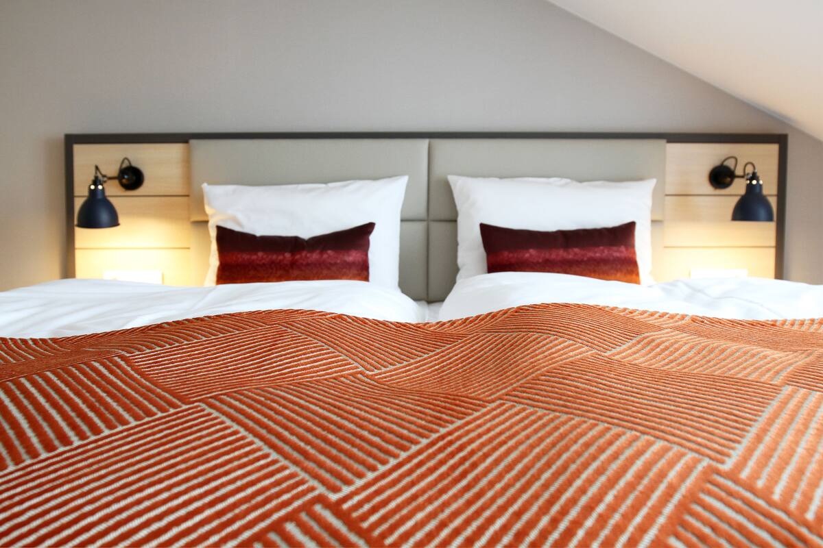 Cama bem arrumada com manta de padrão geométrico, duas almofadas e duas luminárias fixadas na parede.