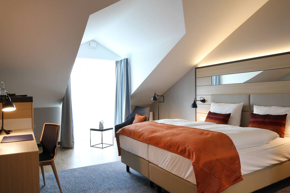 Quarto de hotel moderno com uma cama grande, uma escrivaninha e uma cadeira. Há uma janela com cortinas, um abajur e uma mesa de cabeceira com um candeeiro.
