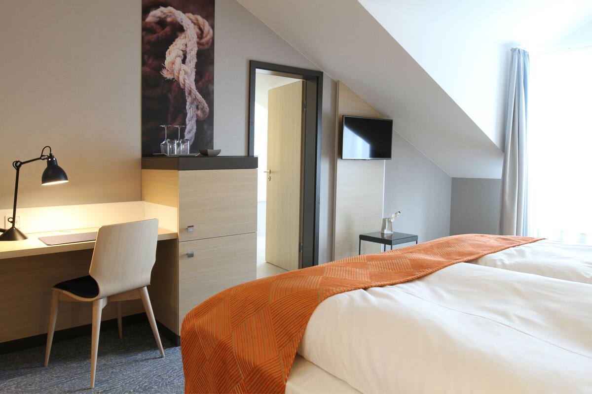 Quarto de hotel com cama, secretária com cadeira, televisão e janela grande.