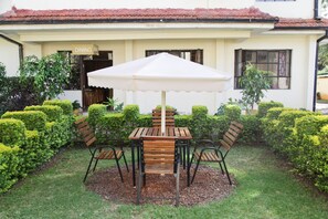 Uma sala de jantar com mesa e cadeiras sob um guarda-sol branco, rodeada por sebes bem cuidadas e um edifício com telhado de azulejos.