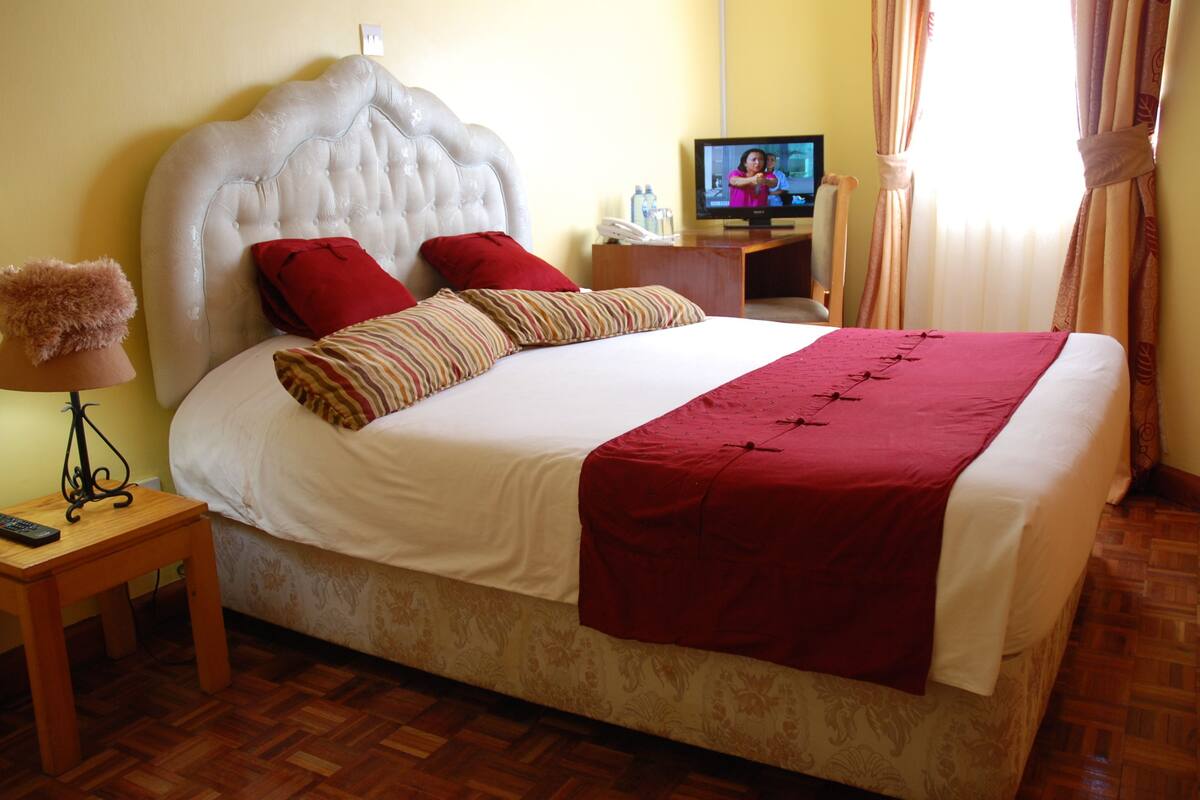 Cama bem arrumada com cabeceira branca, almofadas vermelhas e listradas, e manta vermelha. Um criado-mudo de madeira com abajur e controle remoto, e uma televisão em um suporte de madeira.