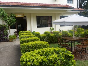 Uma área de refeições com sebes bem aparadas, um espaço coberto para sentar e a entrada de um edifício com a placa 'DINING'.