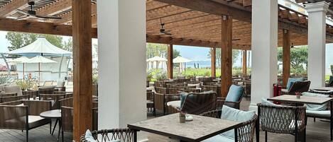 Um pátio ao ar livre com mesas de madeira e cadeiras de vime, coberto por uma pérgola de madeira com pilares brancos.