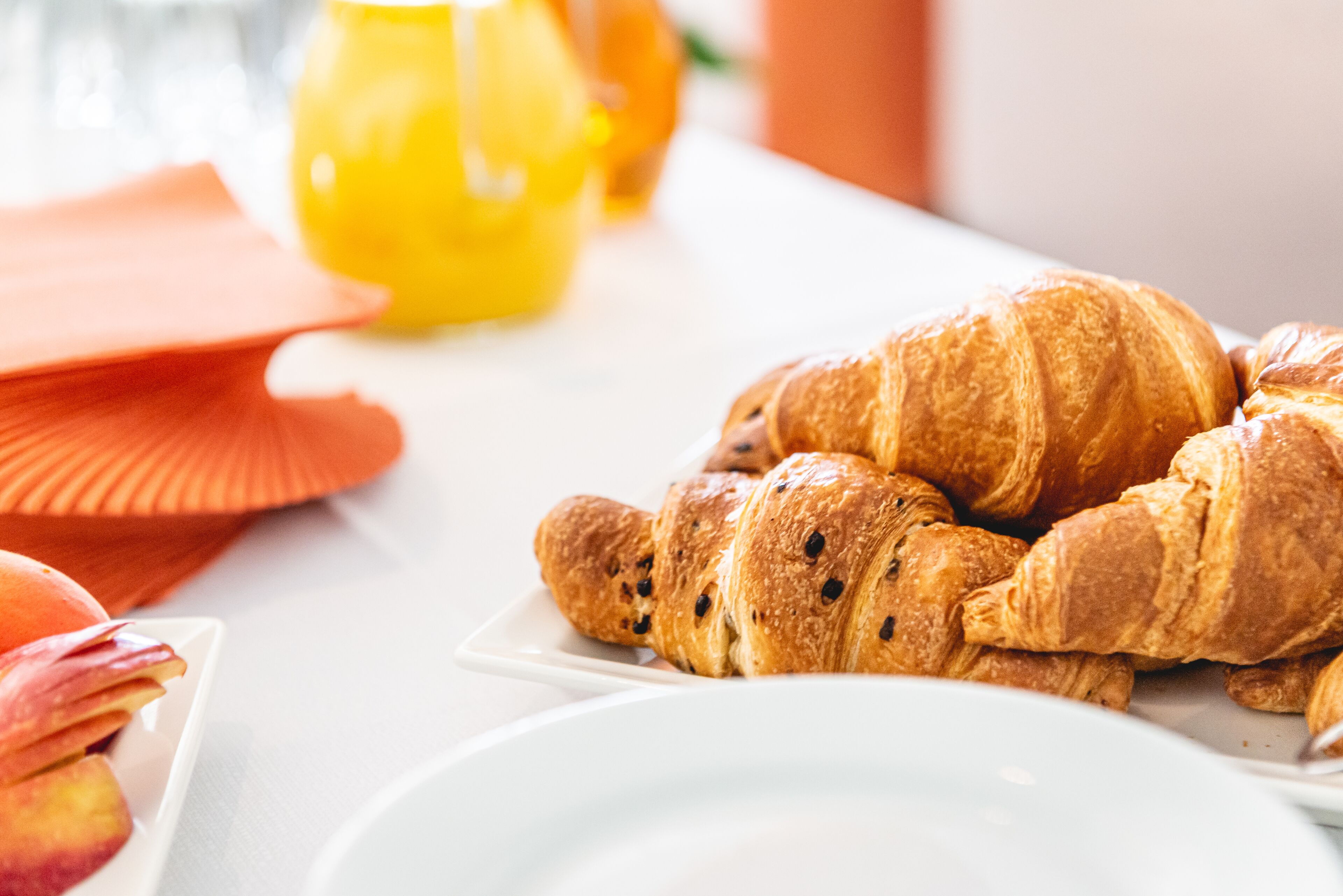 Mesa de café da manhã com croissants em um prato branco, um vaso amarelo e guardanapos laranja.