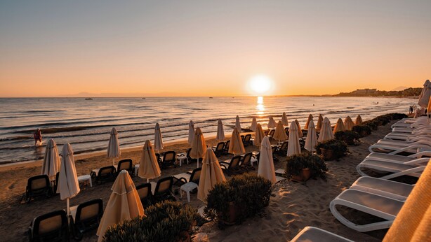 Hotel à beira-mar com fileiras de espreguiçadeiras e guarda-sóis, vista desobstruída do oceano e um pôr do sol ao fundo.