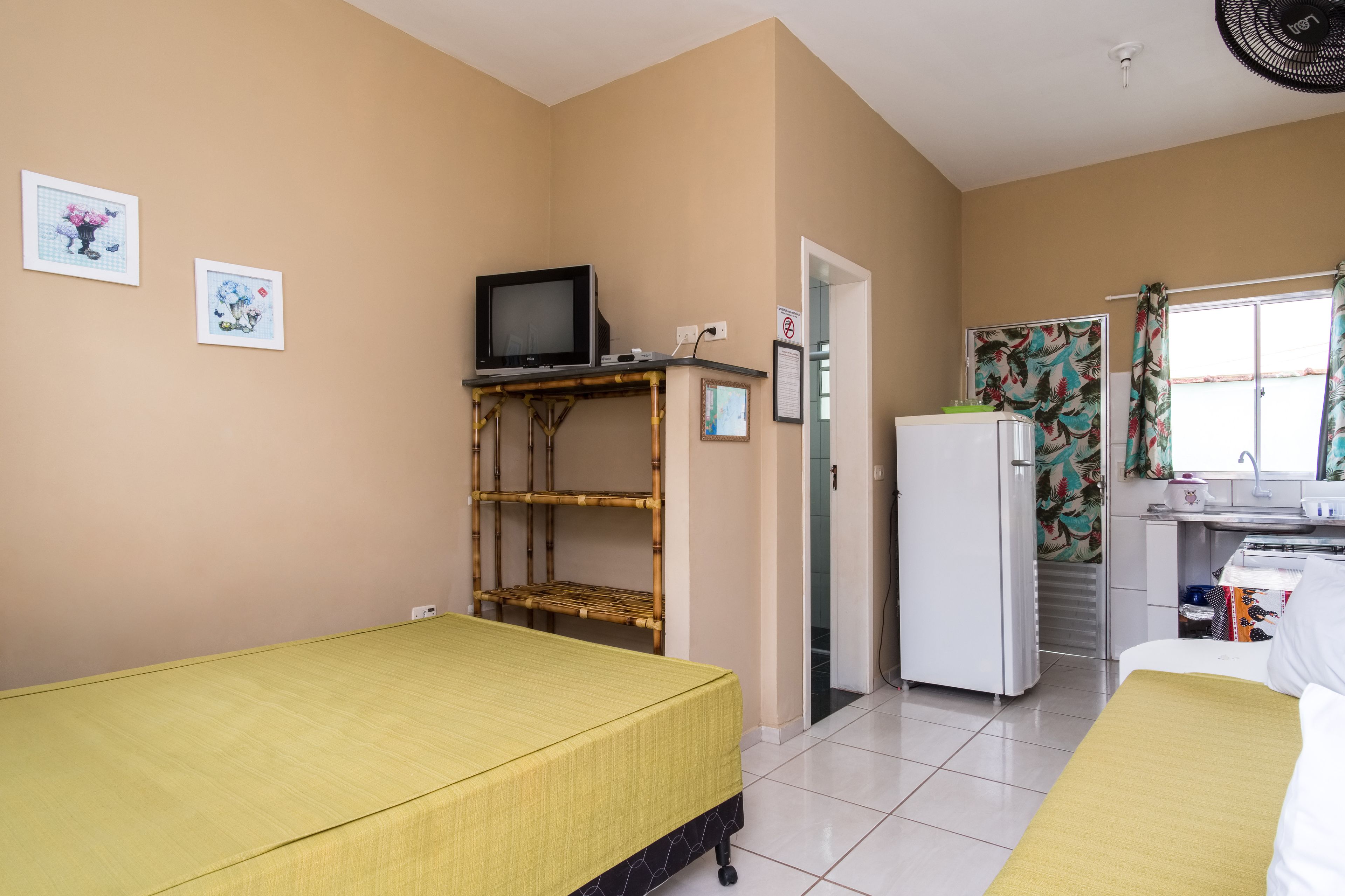 Quarto compacto com cama, uma pequena TV em uma estante de bambu, um refrigerador e uma janela com cortinas.