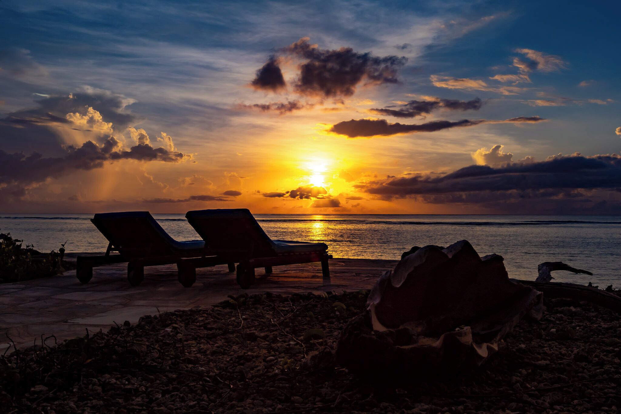 Pôr do sol sobre uma praia, com cadeiras de praia e uma grande formação rochosa.