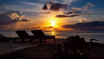 Pôr do sol sobre uma praia, com cadeiras de praia e uma grande formação rochosa.