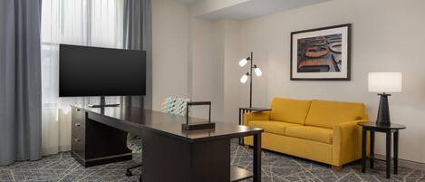 Quarto de hotel com escrivaninha, sofá, televisão, quadro emoldurado e abajur.