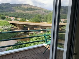 Varanda com vista para uma ponte sobre um rio, cercada por vegetação e colinas.