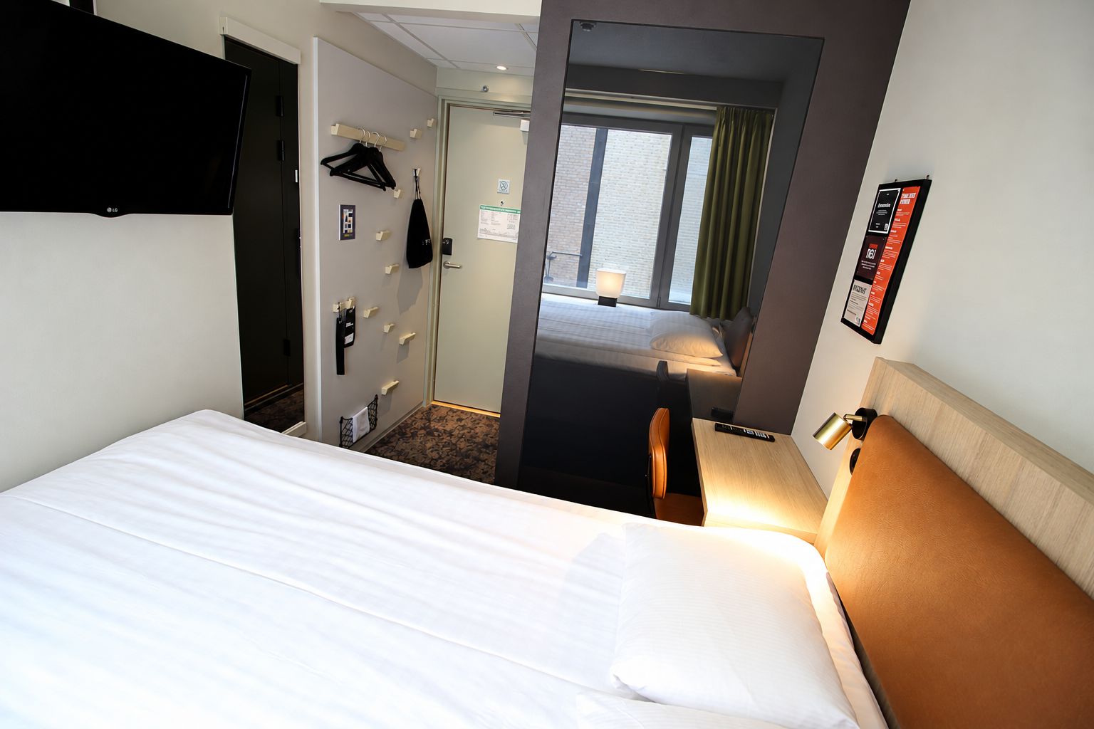 Quarto de hotel com cama, escrivaninha, cadeira, televisão, guarda-roupa e janela com cortinas.