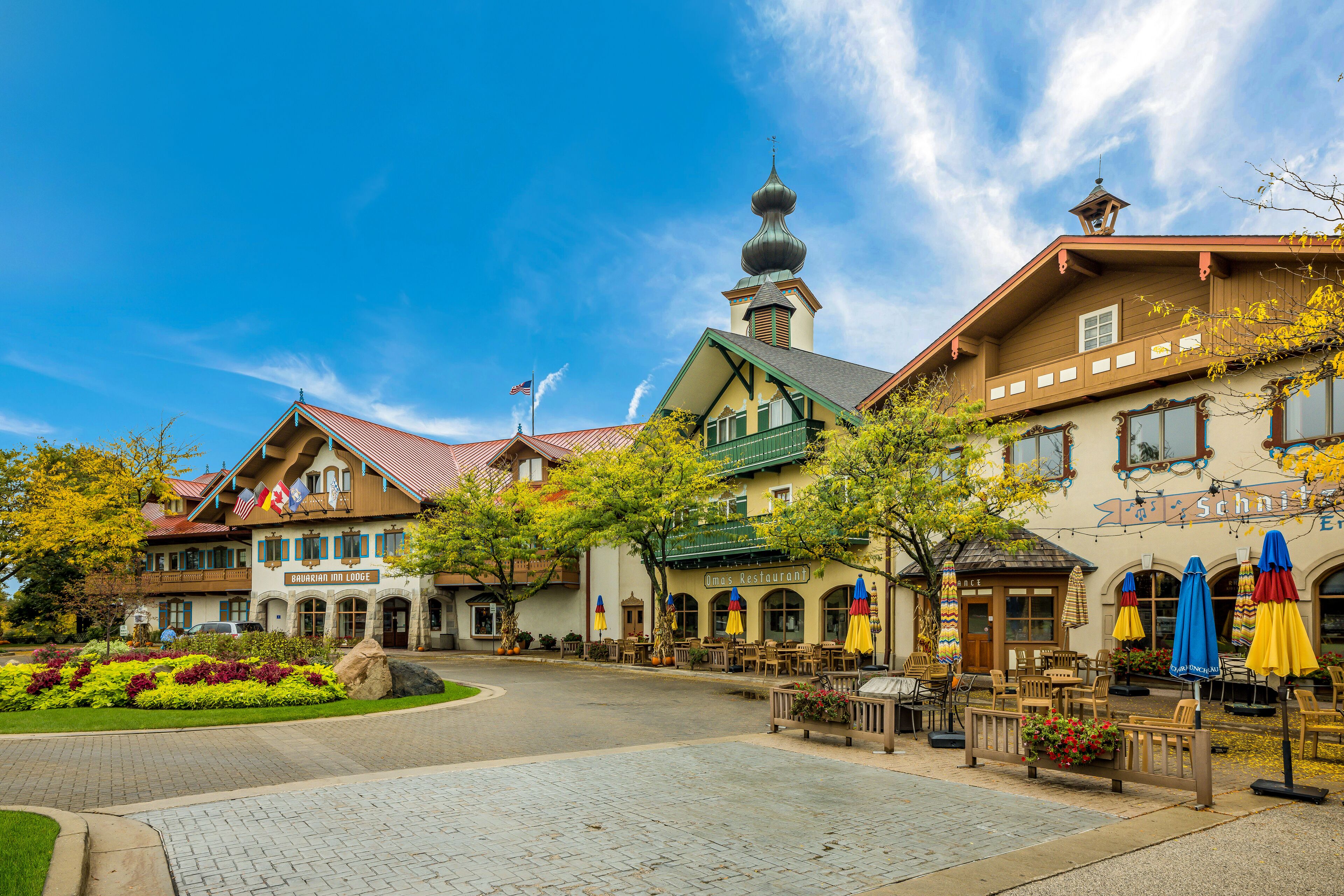 Um edifício de estilo europeu tradicional, com uma torre central, área de estar externa com guarda-sóis e rua de paralelepípedos.