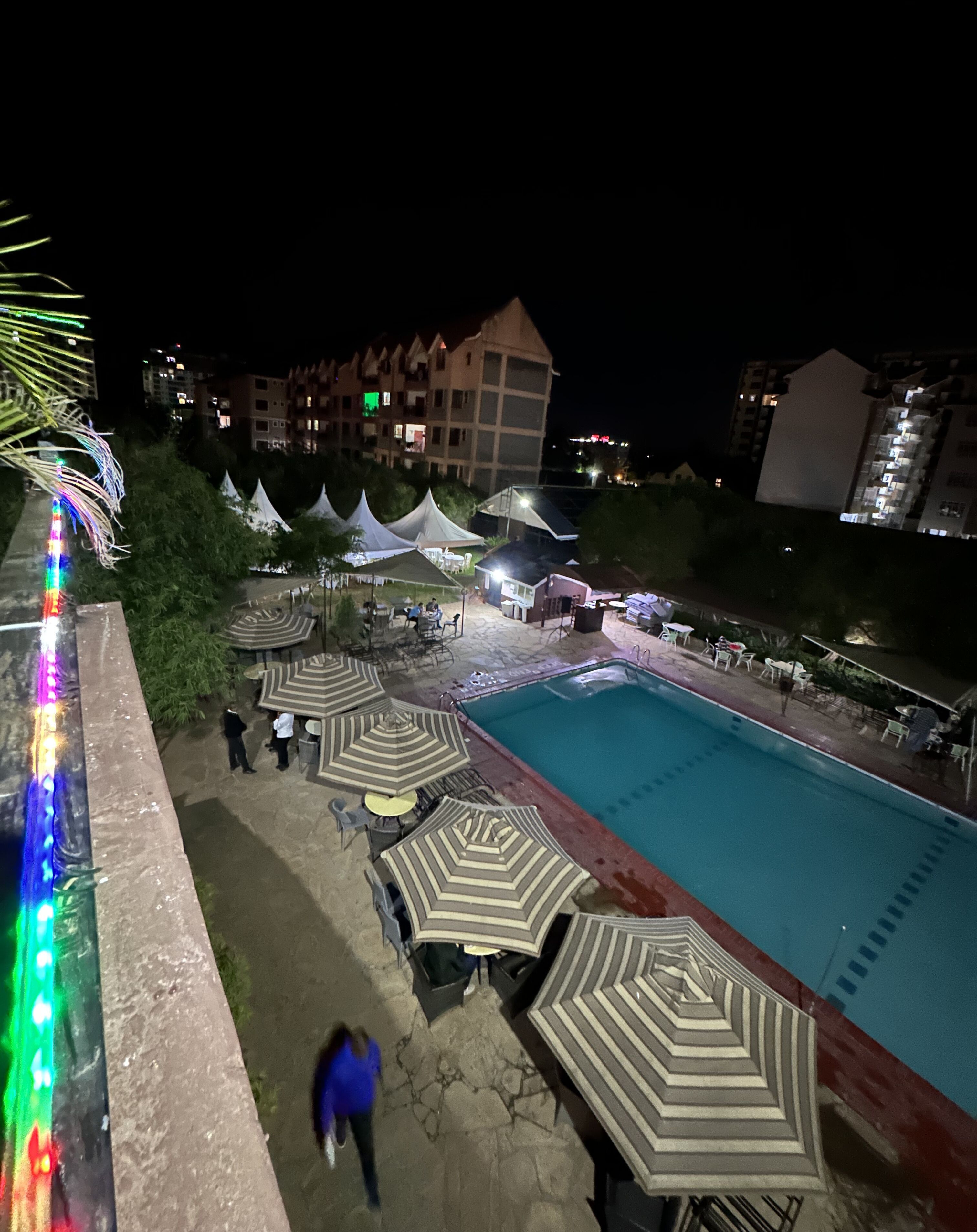 Vista noturna de uma área de piscina de hotel, com guarda-sóis, espreguiçadeiras e uma piscina.