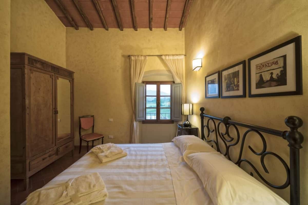 Quarto com cama, guarda-roupa de madeira, cadeira, janela com cortinas e quadros nas paredes.