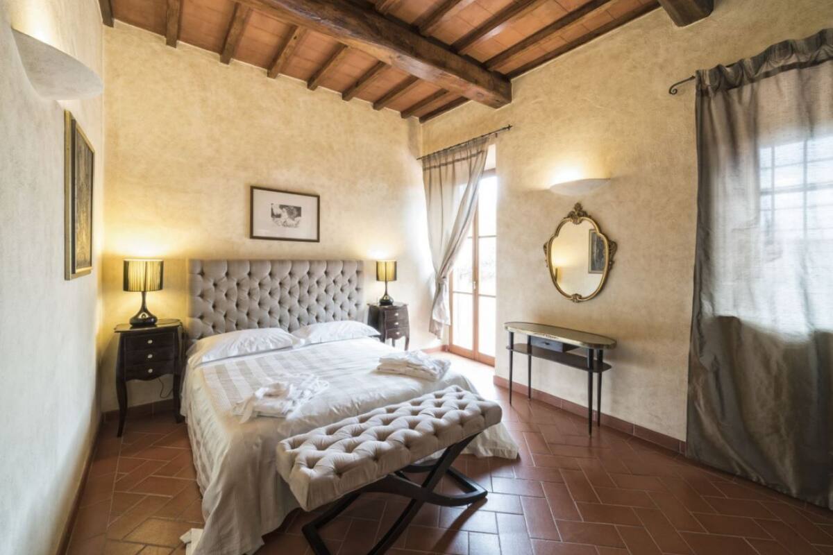 Quarto com cama e cabeceira estofada, luminárias de cabeceira, uma mesinha, um espelho e uma janela com cortinas.