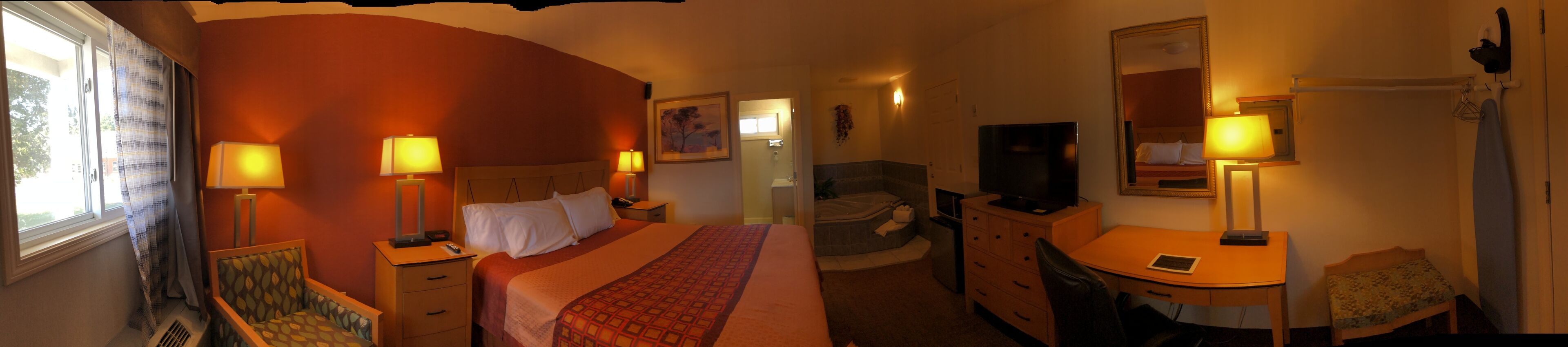Quarto de hotel com cama, mesas de cabeceira com abajures, escrivaninha, cadeira, televis ão, espelho e banheiro com banheira e vaso sanitário.