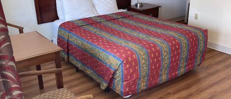 Quarto de hotel com cama de madeira, uma escrivaninha, uma cadeira e uma mesa de cabeceira.