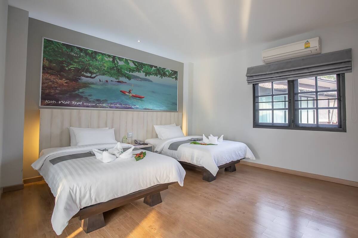 Quarto de hotel com duas camas, um quadro grande de uma paisagem tropical, um aparelho de ar condicionado e uma janela com cortinas.