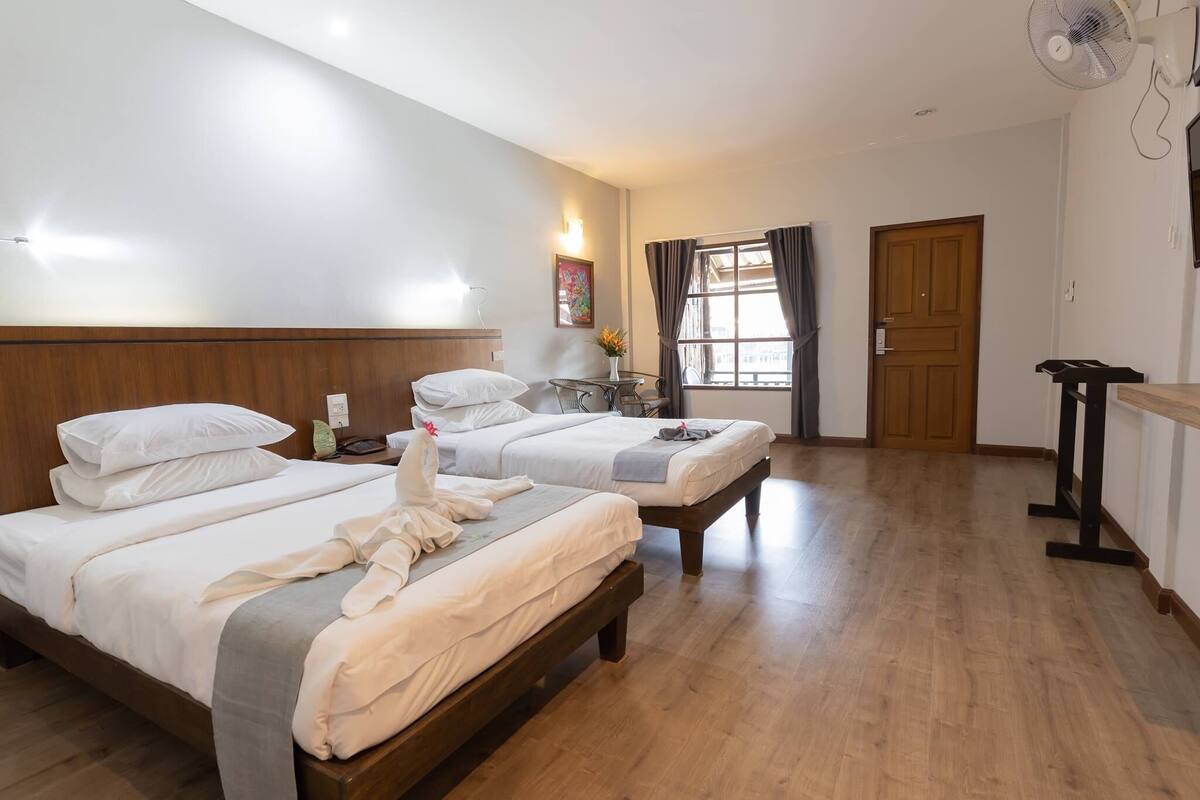 Quarto de hotel com duas camas, uma mesinha de cabeceira, uma pequena mesa com um vaso, uma janela com cortinas e uma porta.