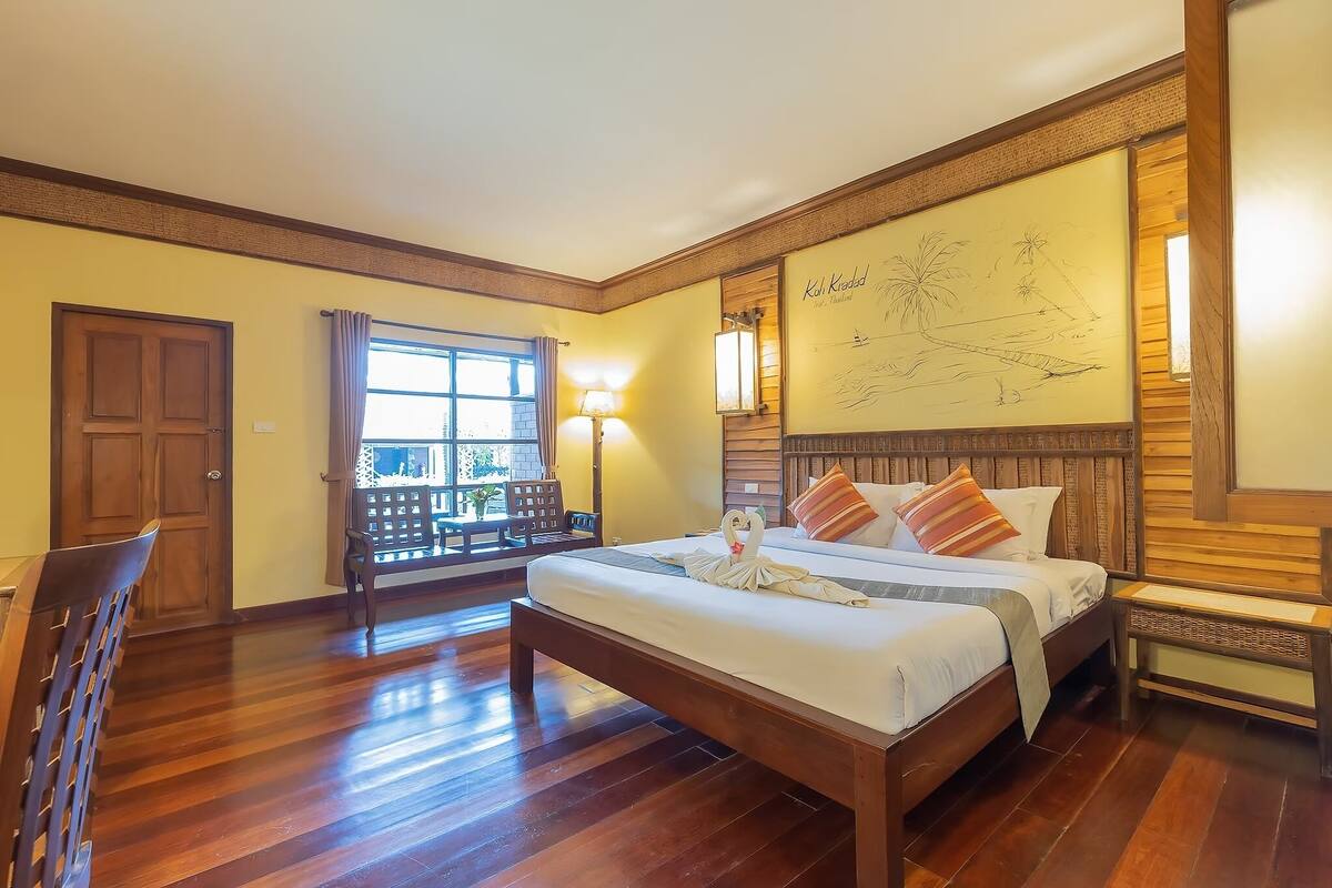 Quarto de hotel com cama de madeira, uma janela grande com cortinas, luminária na parede e uma obra de arte emoldurada.