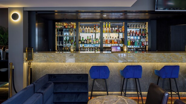 Uma área de bar com bancada de mármore, banquetas azuis e diversas garrafas de bebidas alcoólicas.