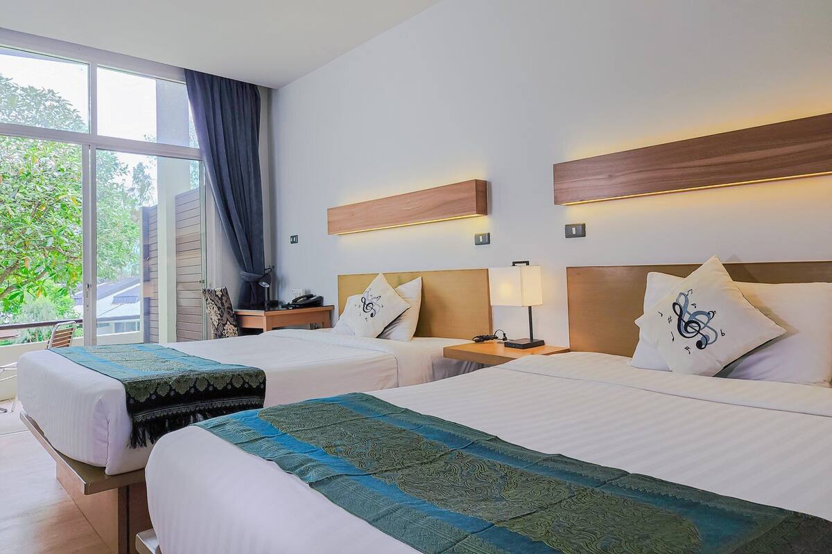Quarto de hotel com duas camas, uma janela grande com cortinas, uma mesinha de cabeceira com abajur e um ponto de luz fixado na parede.