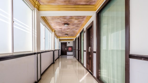 Um corredor comprido e estreito, com chão de azulejos, janelas amplas de um lado e portas do outro.