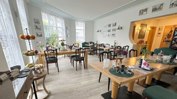 Uma área de refeições com várias mesas e cadeiras, um buffet com frutas e pastelaria, e uma parede decorada com quadros.