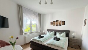 Quarto com uma cama grande, mesa de cabeceira, uma cadeira, janela com cortinas e televisão fixada na parede.