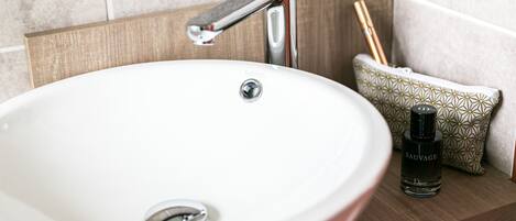 Uma pia de banheiro branca com torneira cromada, um frasco de perfume Sauvage e uma almofada estampada em uma prateleira de madeira.