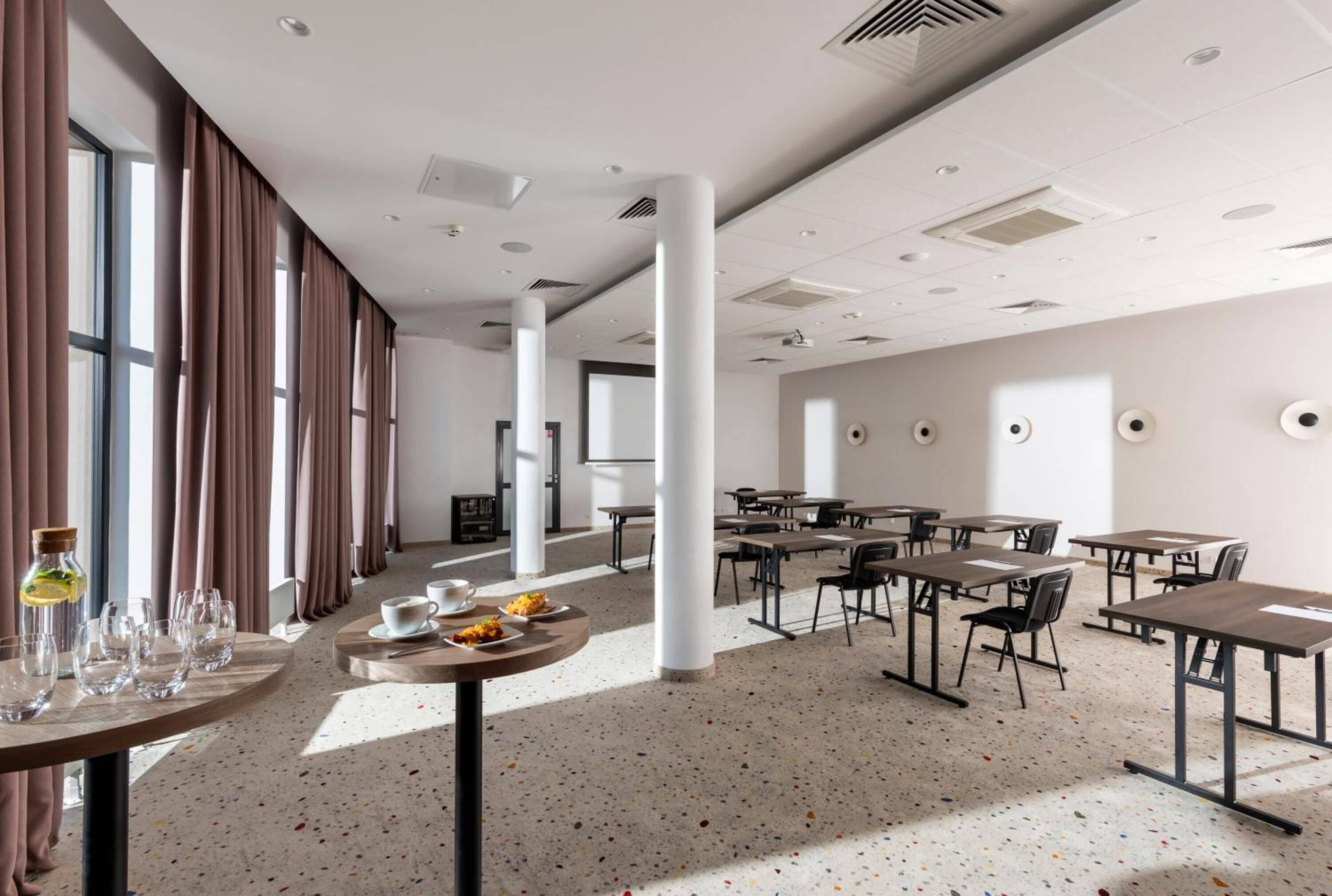 Ampla sala de jantar de hotel com janelas grandes, várias mesas e cadeiras.