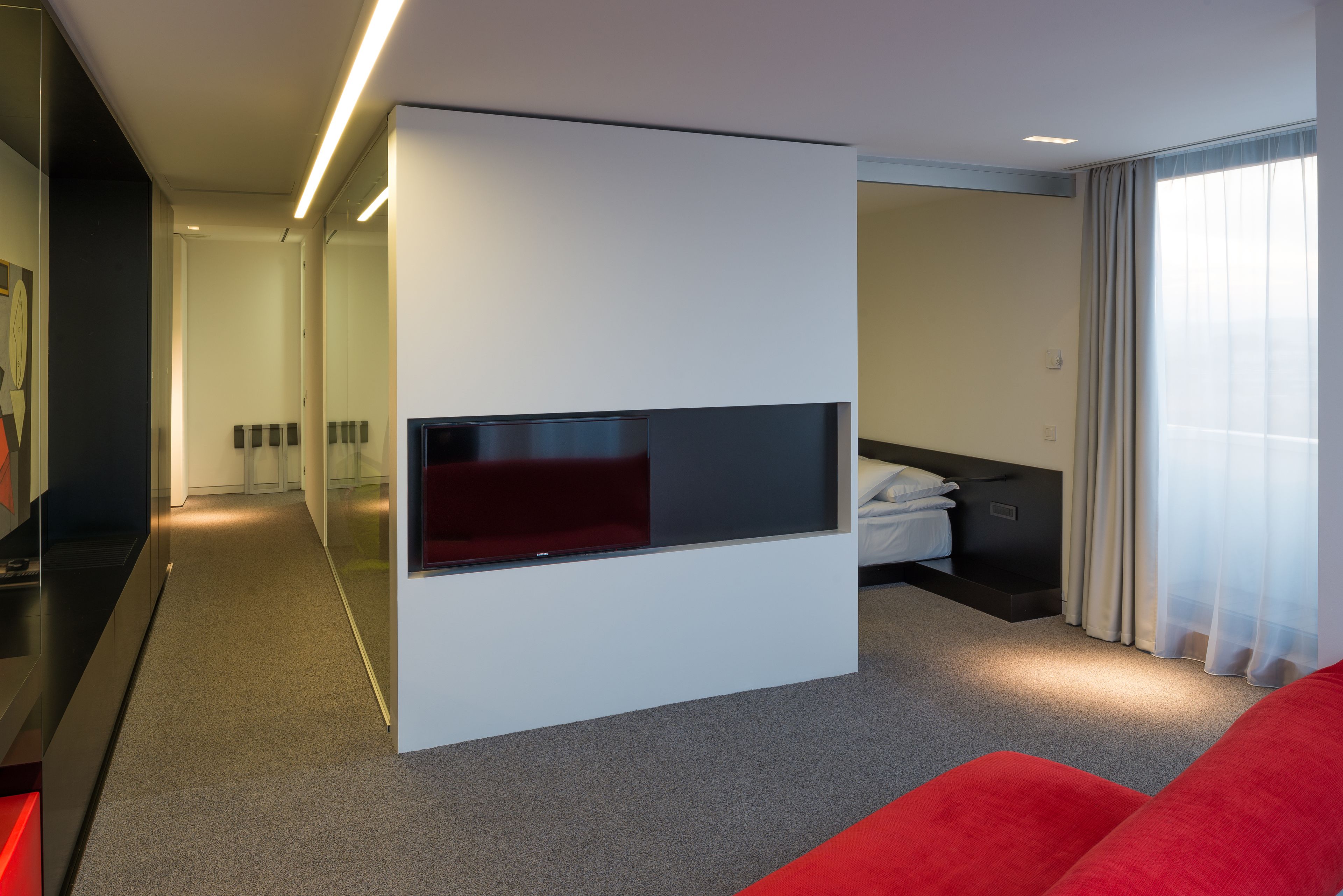 Quarto de hotel moderno com uma televisão de tela plana grande, um sofá vermelho, uma cama com lençóis brancos e uma janela ampla com cortinas leves.