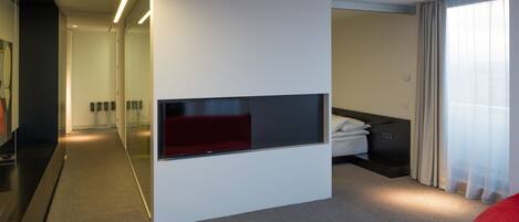 Quarto de hotel moderno com uma televisão de tela plana grande, um sofá vermelho, uma cama com lençóis brancos e uma janela ampla com cortinas leves.