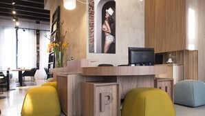 Uma área de recepção moderna com uma mesa de madeira, um computador e um vaso de flores. Há dois pufes amarelos e um azul.