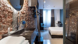 Quarto de hotel moderno com parede de destaque em pedra, uma cama grande, uma cama menor, uma escrivaninha com computador e um banheiro com pia e espelho.