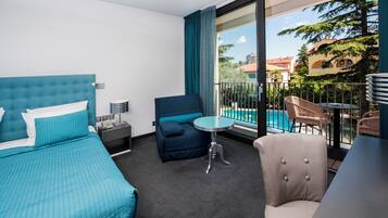 Quarto de hotel com cama, sofá, mesa pequena e varanda com vista para a piscina e edifícios.