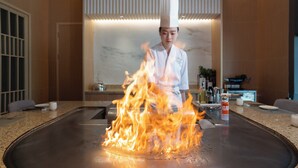 Um chef demonstra culinária com fogo em uma cozinha moderna com painel de mármore e elementos de design contemporâneos.