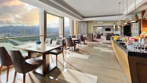 Uma área de refeições moderna em hotel com vista panorâmica da cidade, um bar com frutas e copos, e um espaço de estar aconchegante.