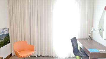 Quarto de hotel com uma janela grande, uma escrivaninha com cadeira e uma cama.