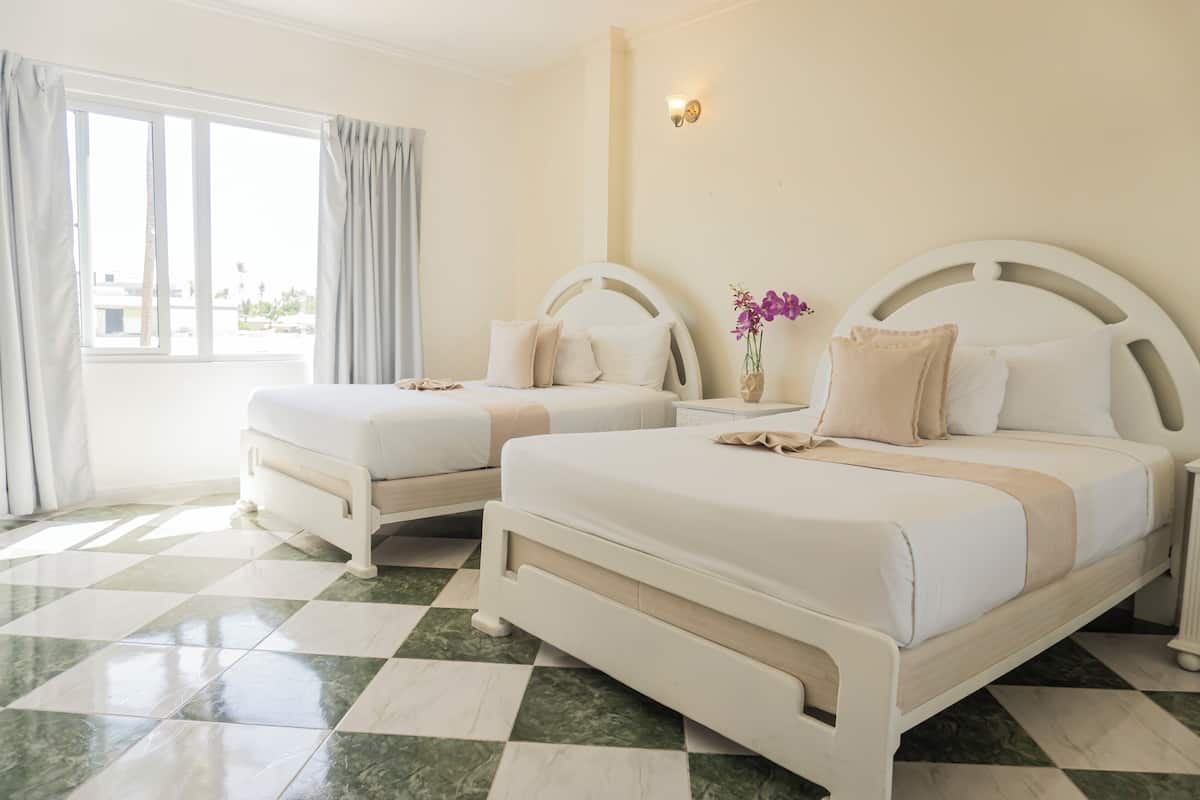 Quarto de hotel com duas camas, piso de azulejos quadriculados, uma janela com cortinas e luminária fixada na parede.