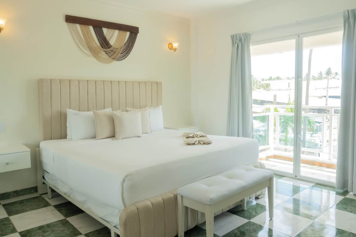 Quarto de hotel com uma cama grande, um banco, uma mesinha de cabeceira, uma luminária na parede e vista para o exterior através de uma porta de correr.